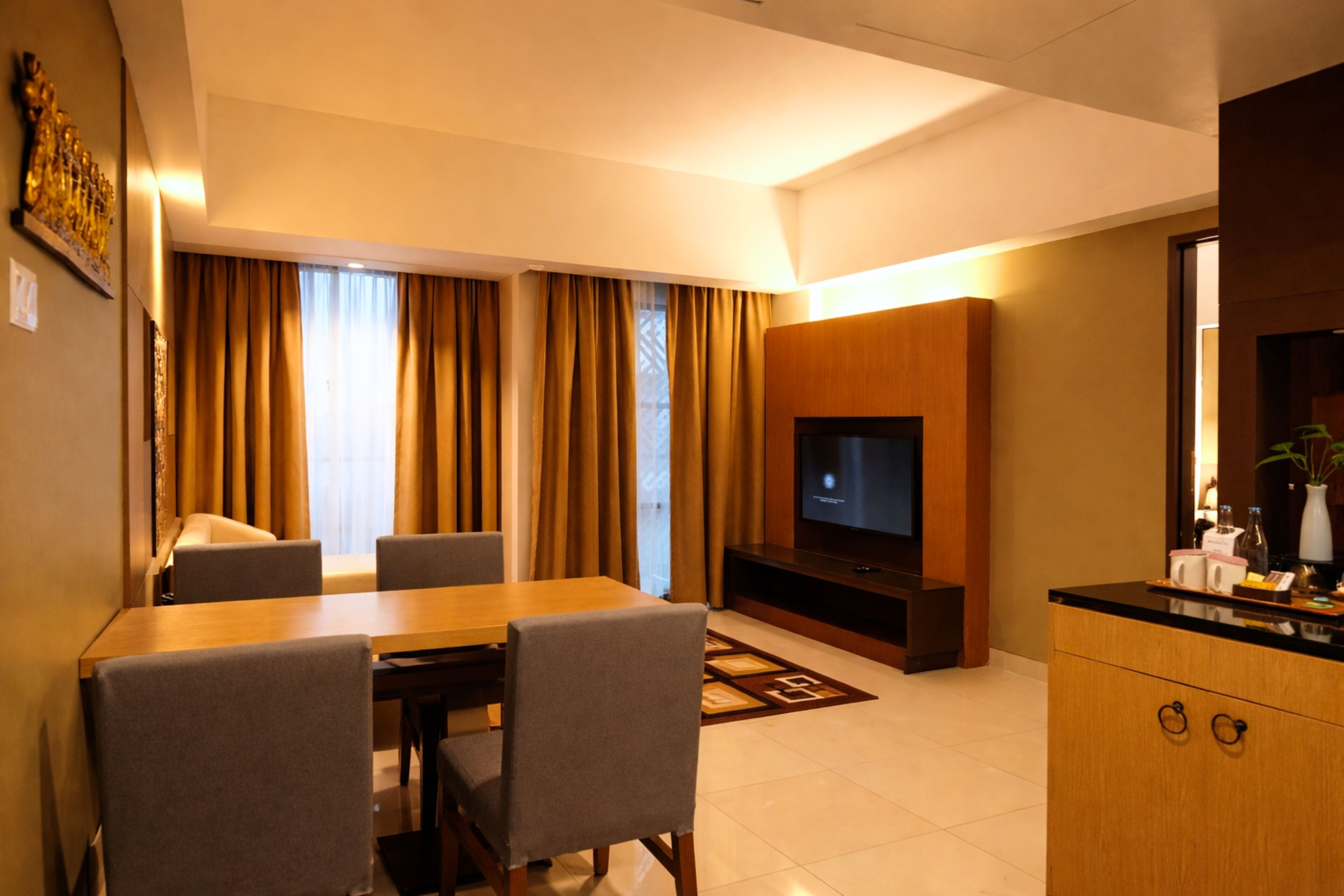 Quarto de hotel com área de refeições, televisão, armário de madeira e janela com cortinas.