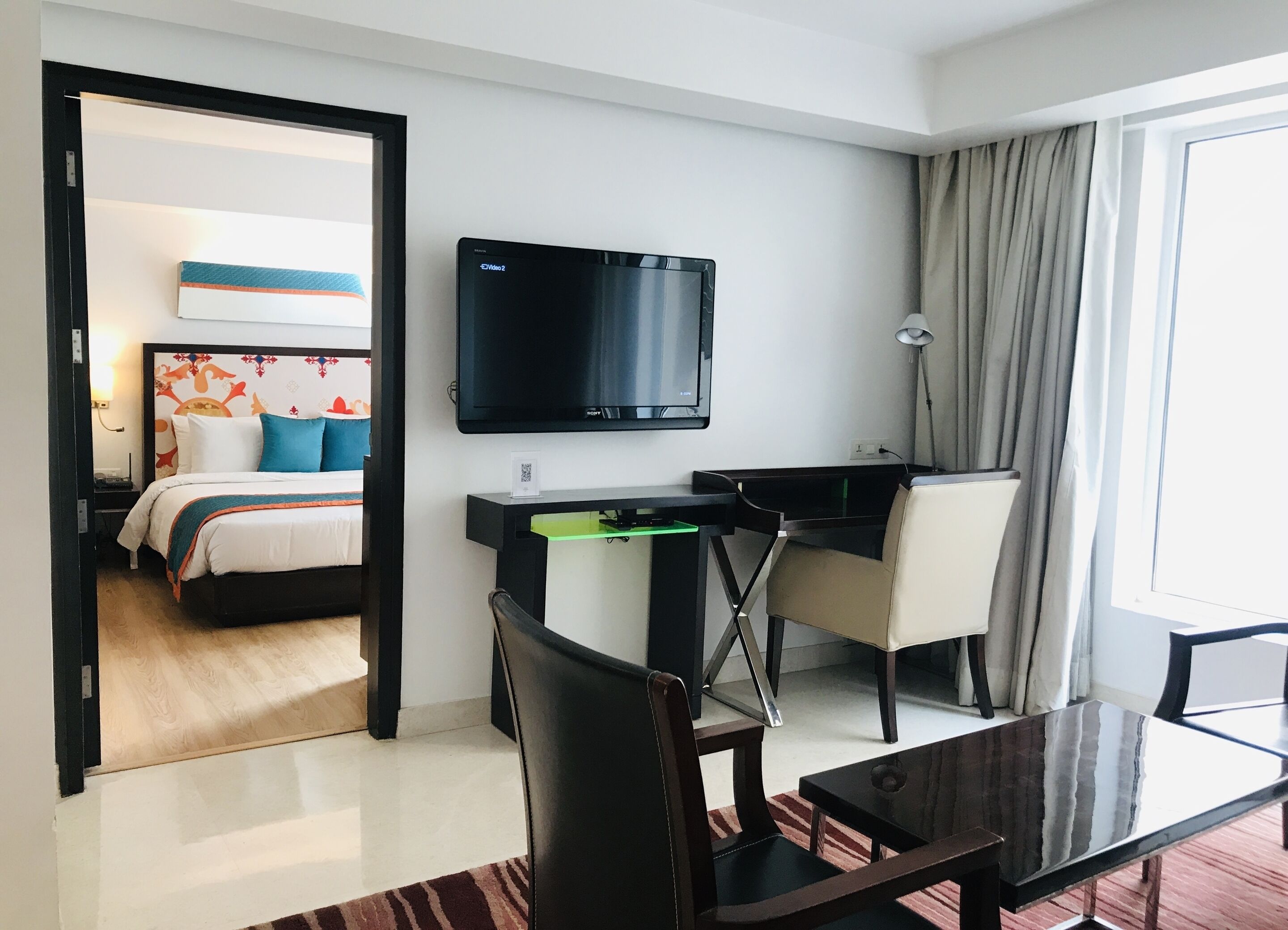 Quarto de hotel com cama, escrivaninha, cadeira, televisão e janela com cortinas.