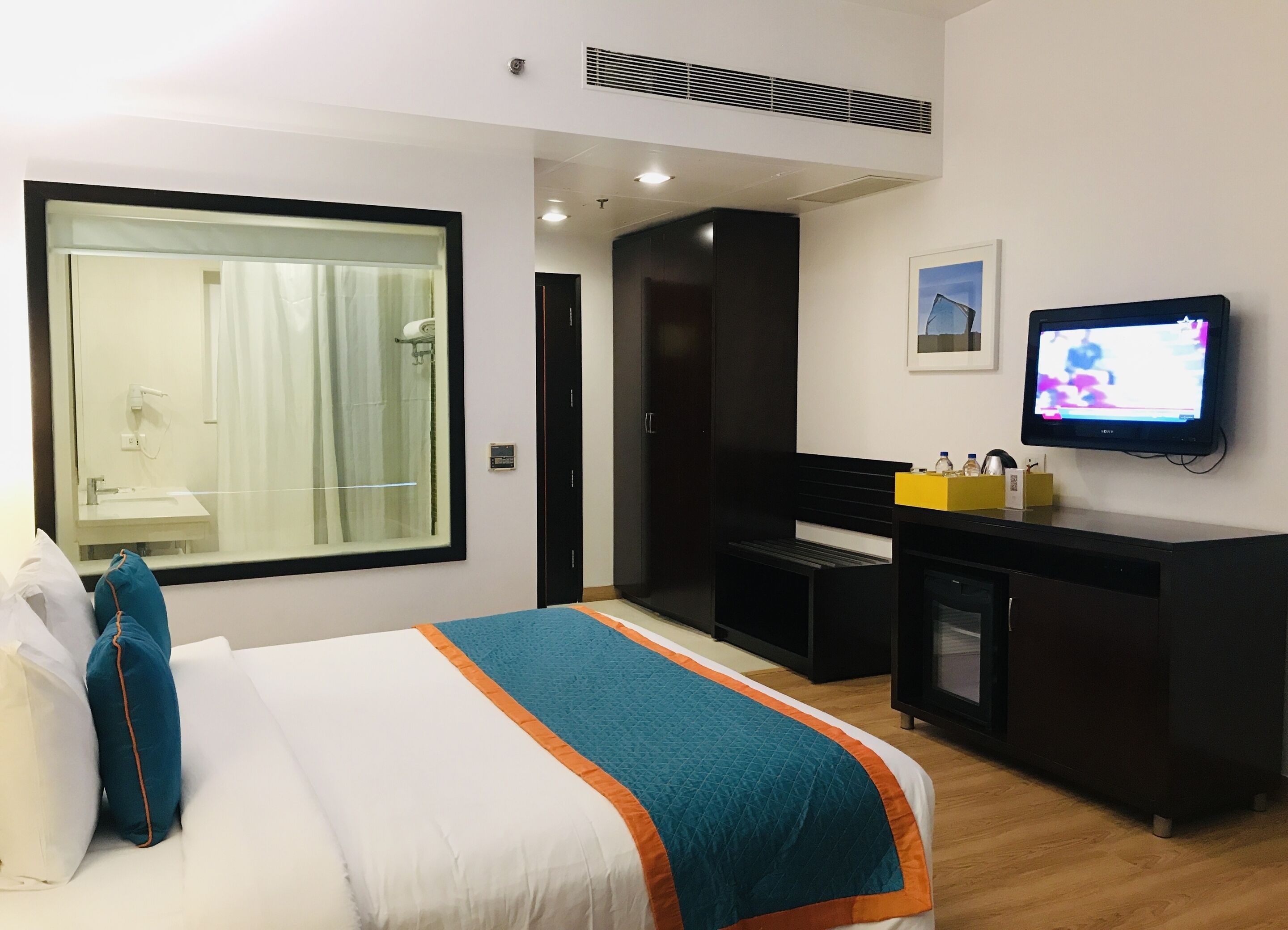 Quarto de hotel com uma cama grande, uma escrivaninha, televisão e banheiro visível por meio de um painel de vidro.