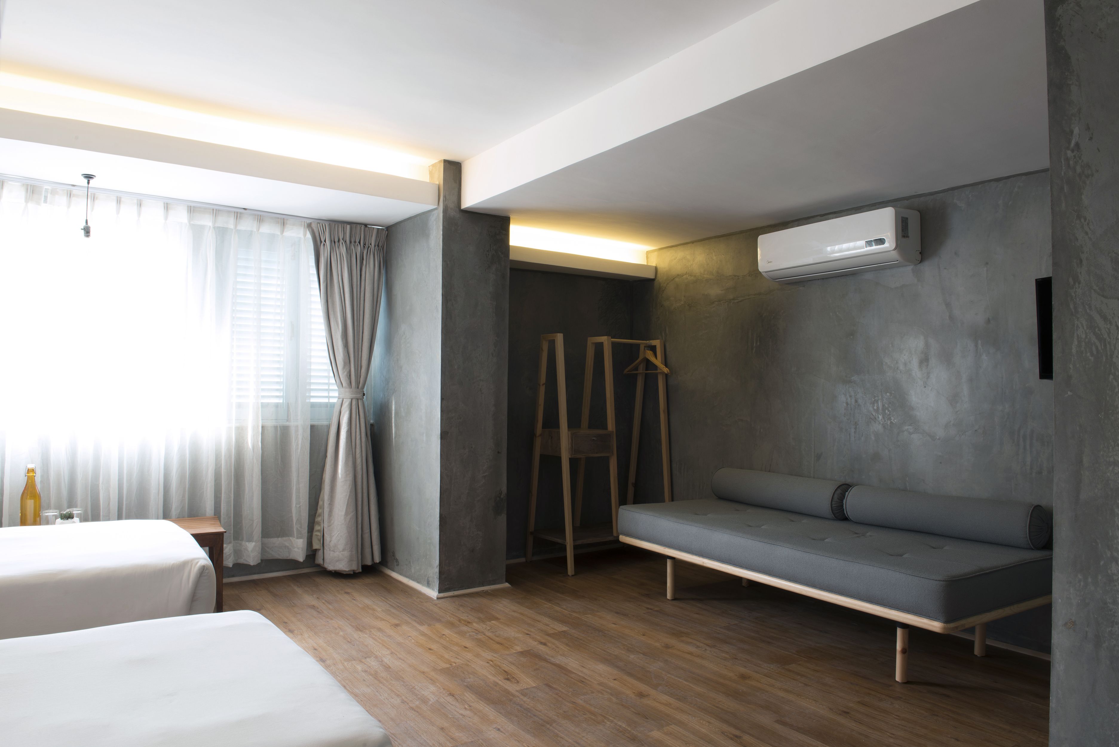 Quarto de hotel moderno com piso de madeira, um sofá cinza, uma cama, uma mesinha e uma janela com cortinas.