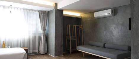 Quarto de hotel moderno com piso de madeira, um sofá cinza, uma cama, uma mesinha e uma janela com cortinas.
