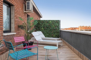 Terraço no telhado com parede de tijolos, parede verde com flores, duas cadeiras de descanso, uma mesa pequena e um vaso com planta.