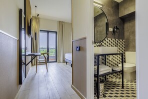 Quarto de hotel moderno com varanda, janela grande, cama, escrivaninha e banheiro com piso xadrez e vaso sanitário.