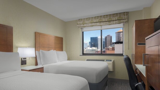 Quarto de hotel com duas camas, uma escrivaninha, uma cadeira e uma janela com vista para a cidade.