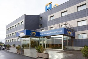 Um hotel moderno com fachada azul e cinzenta, exibindo um logo 'H' em destaque e uma placa indicando que possui restaurante e café.