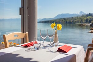Mesa posta com taças de vinho, um vaso com flores amarelas e um guardanapo vermelho, com vista para um lago e montanhas ao fundo.