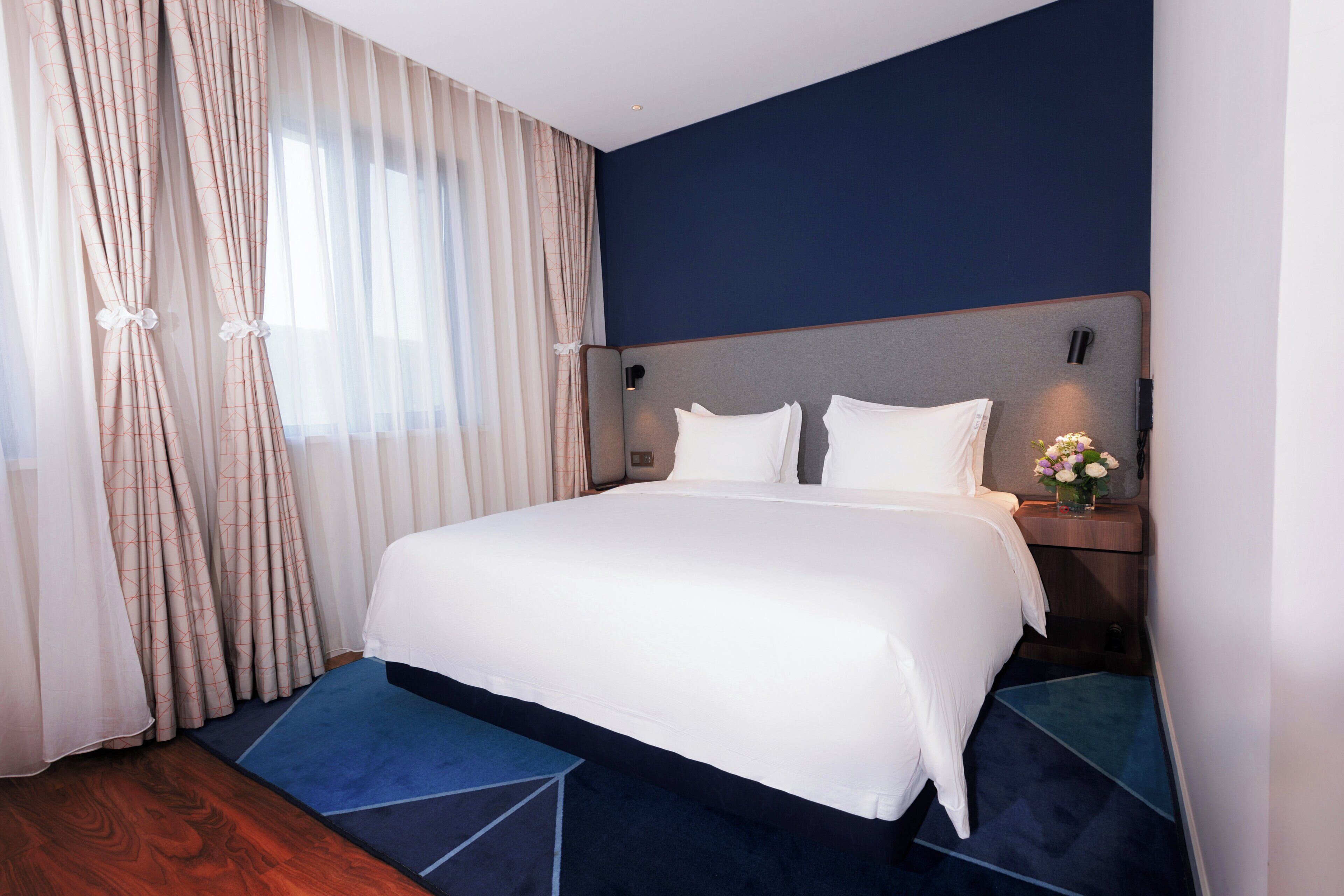 Quarto de hotel com uma cama grande, mesa de cabeceira com um vaso de flores e vista para o exterior através de cortinas leves.