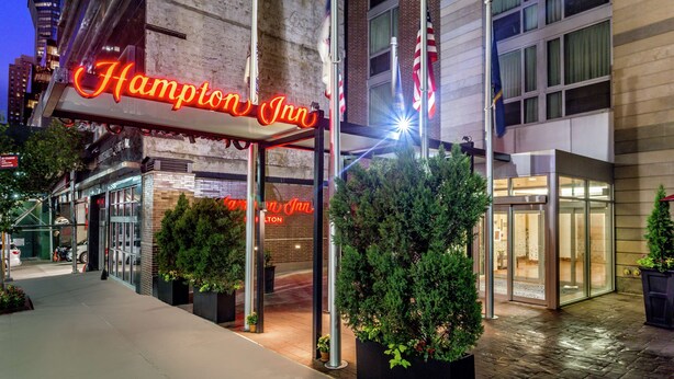 Exterior do Hampton Inn, com letreiro de neon, bandeiras americanas e fachada de tijolos.