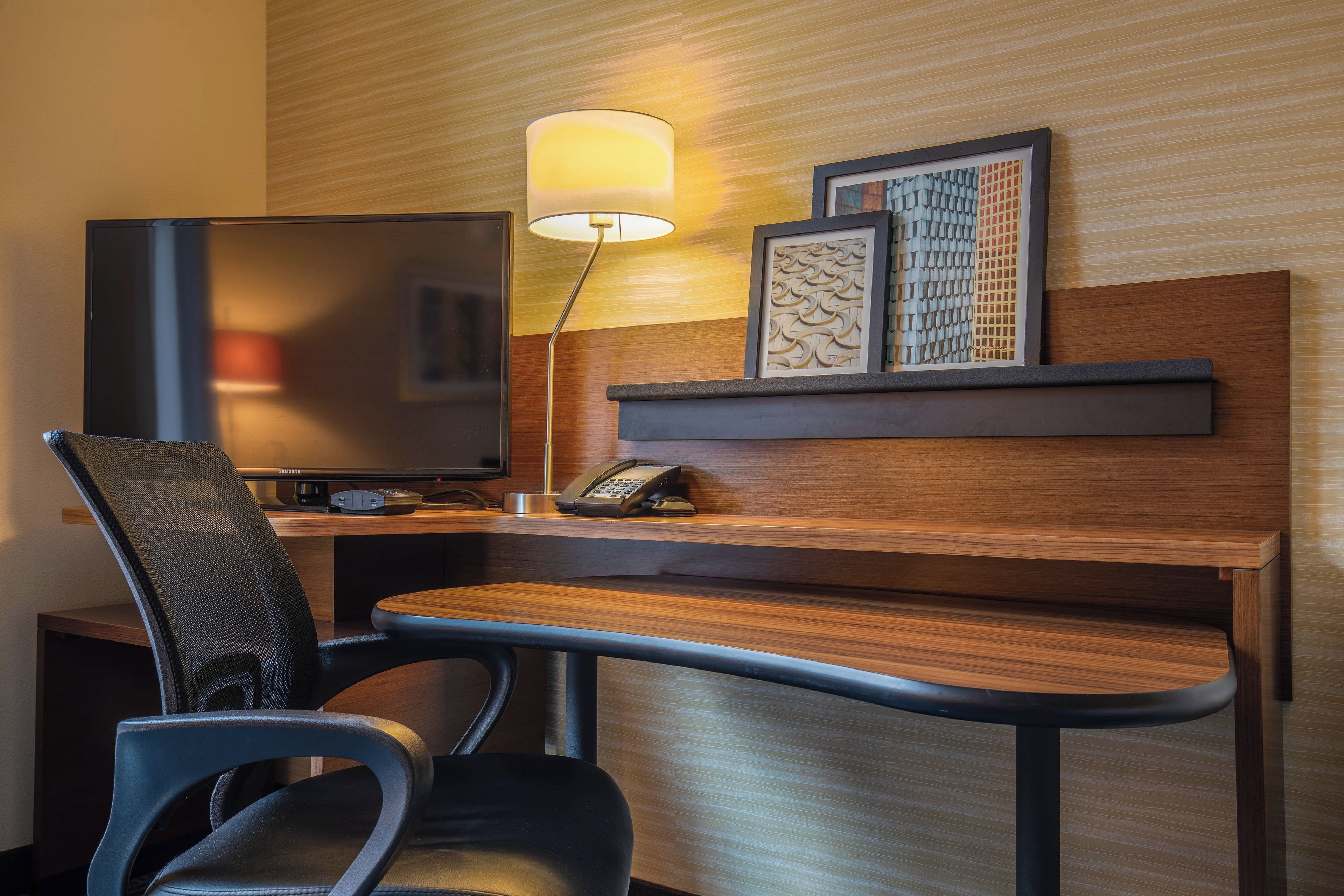 Quarto de hotel com escrivaninha, cadeira, televisão, abajur e telefone.