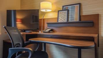 Quarto de hotel com escrivaninha, cadeira, televisão, luminária e telefone.