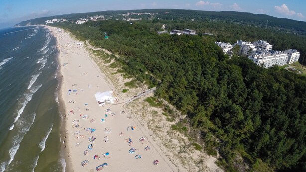 Vista aérea de uma praia com vários guarda-sóis e pessoas, cercada por uma floresta densa e edifícios ao fundo.