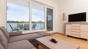 Uma sala de estar moderna com uma grande porta de vidro deslizante que dá para uma varanda com vista para o mar. Há uma televisão de tela plana na parede e uma mesa de centro de madeira com duas xícaras.