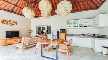 Ampla sala de estar com mesa e cadeiras de madeira, televisão e cozinha com armários brancos e piso ladrilhado.