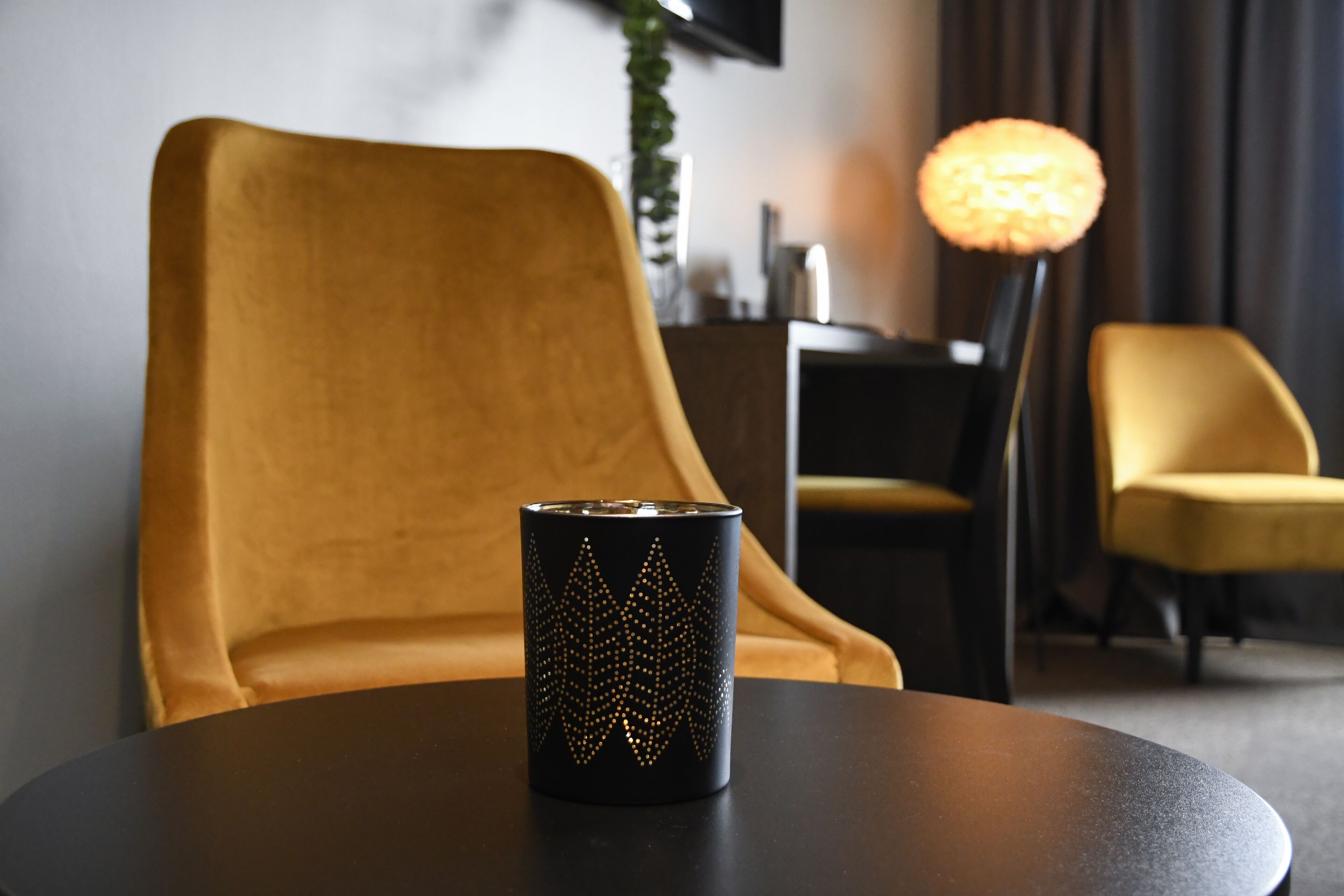 Quarto com mesa preta, caneca preta com padrão dourado, poltrona cor mostarda e abajur com luz esférica.