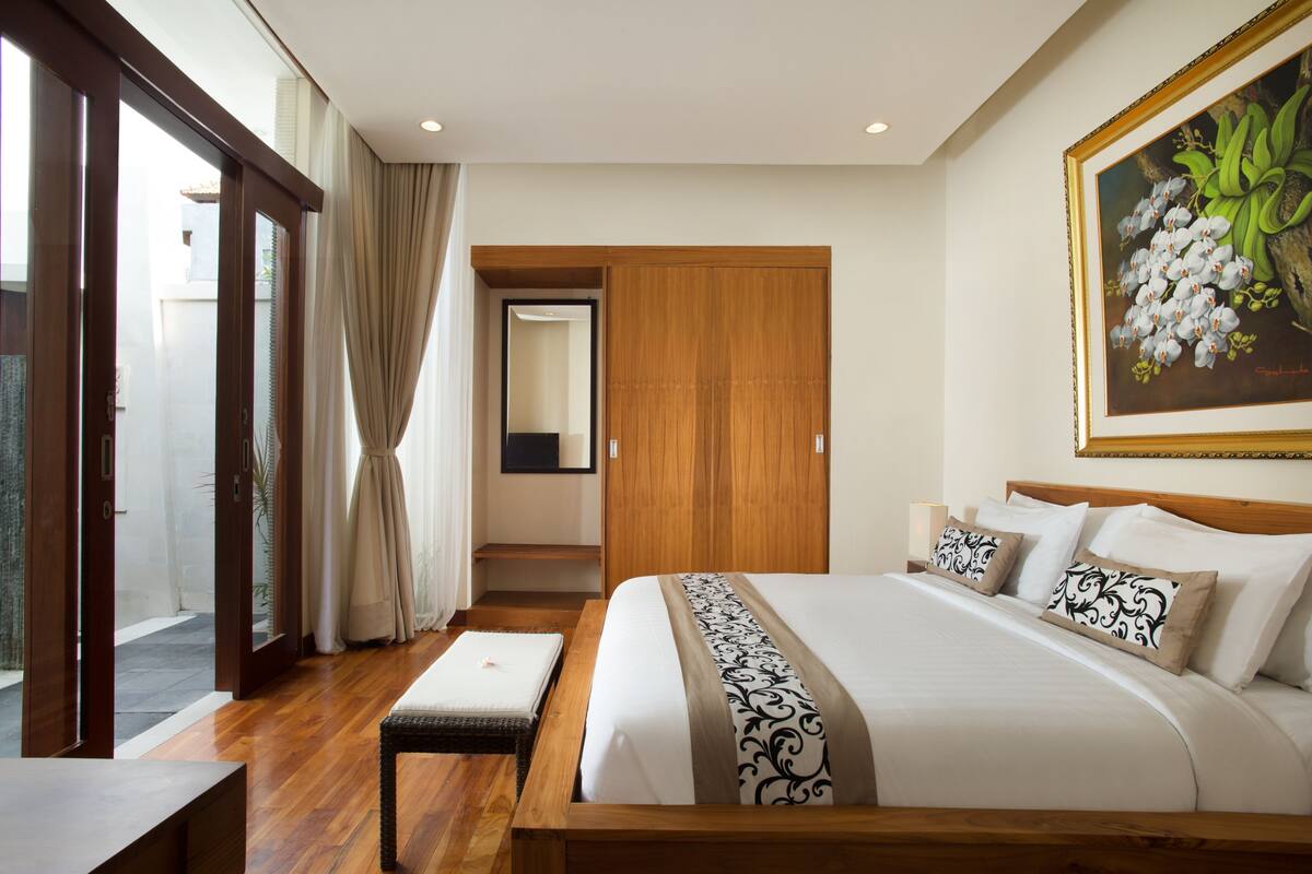 Quarto de hotel com uma cama grande, um banco de madeira, uma porta de vidro deslizante que dá para uma varanda e um quadro de flores na parede.