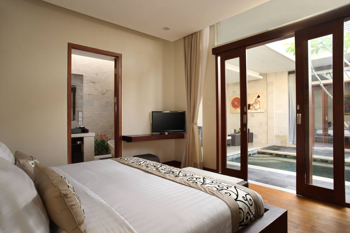 Quarto de hotel com uma cama grande, televisão, escrivaninha e vista para a piscina e área externa.