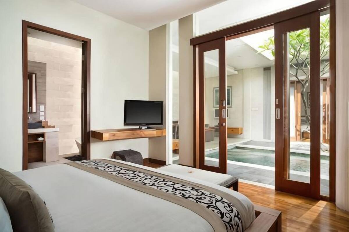 Quarto com cama, televisão, escrivaninha e porta de vidro que dá para uma área de piscina.