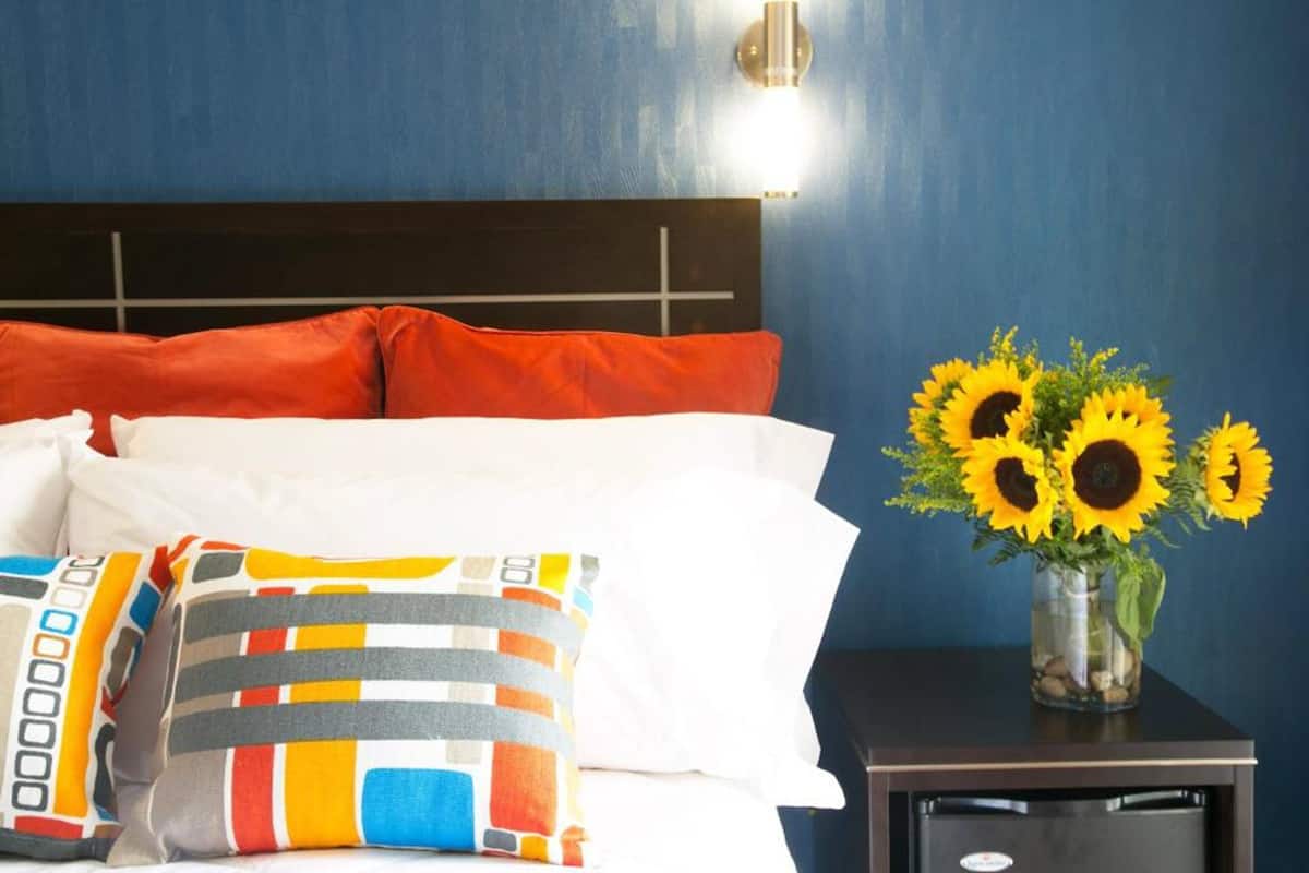 Cama bem arrumada com travesseiros brancos e laranja, um travesseiro listrado colorido e um vaso de girassóis em uma mesa de cabeceira.