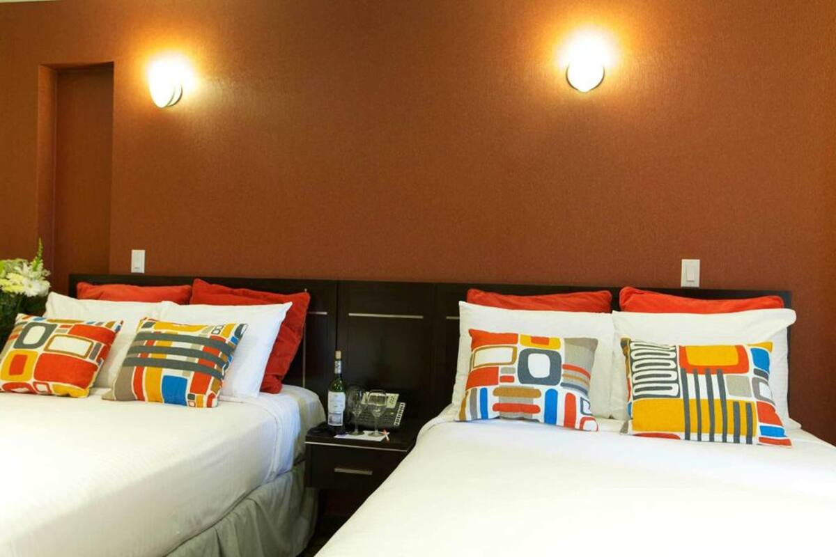 Quarto de hotel com duas camas, cada uma com almofadas coloridas estampadas, mesa de cabeceira com garrafa e telefone, e duas luminárias fixadas na parede.