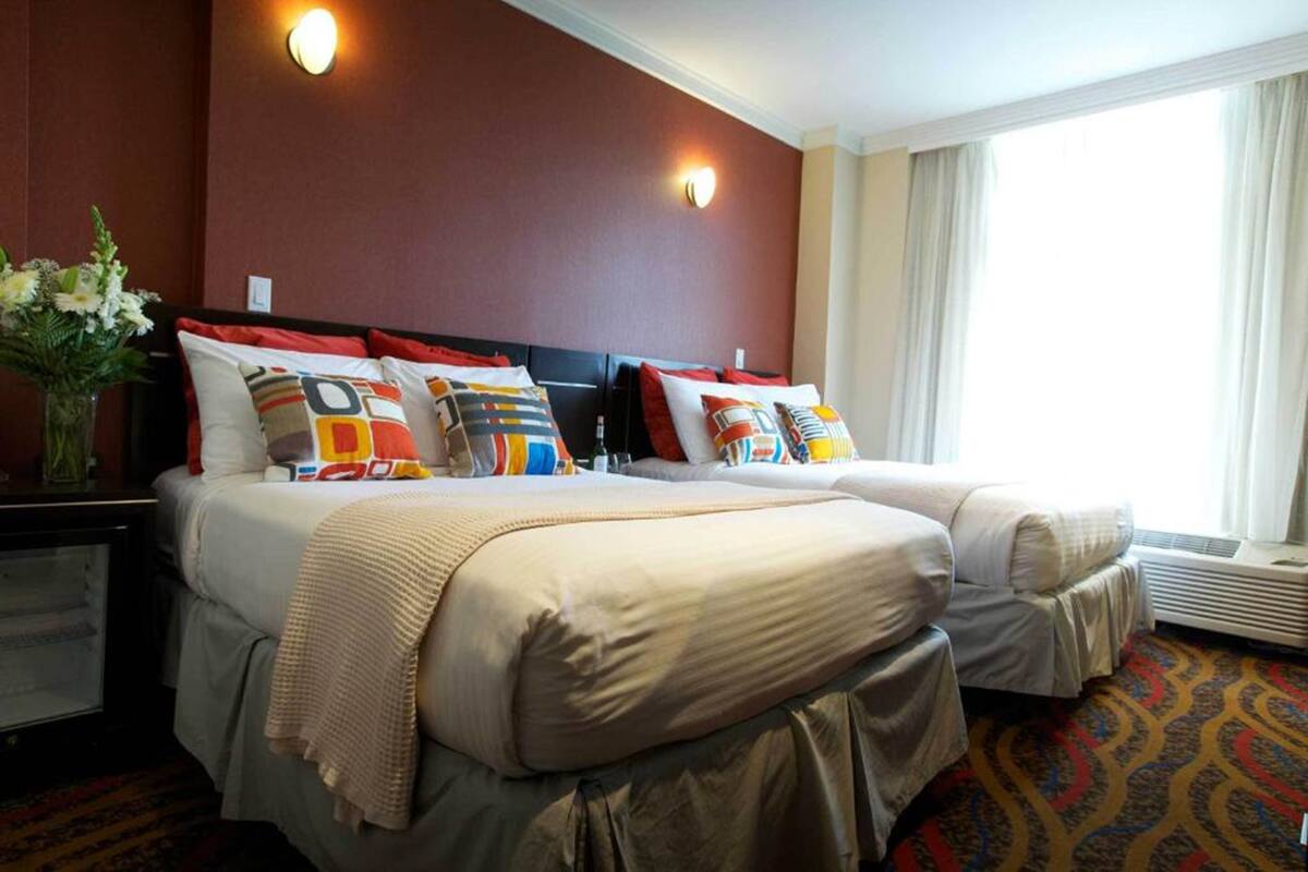 Quarto de hotel com duas camas, uma janela grande com cortinas, uma luminária na parede e um vaso decorativo na mesa de cabeceira.
