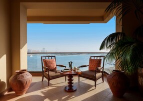 Uma varanda com duas cadeiras de vime, uma mesa redonda pequena e dois vasos grandes. Dá-se para o mar e para o horizonte urbano.