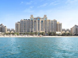 Um grande hotel com vários andares e uma torre central, localizado à beira de uma praia com palmeiras e guarda-sóis.