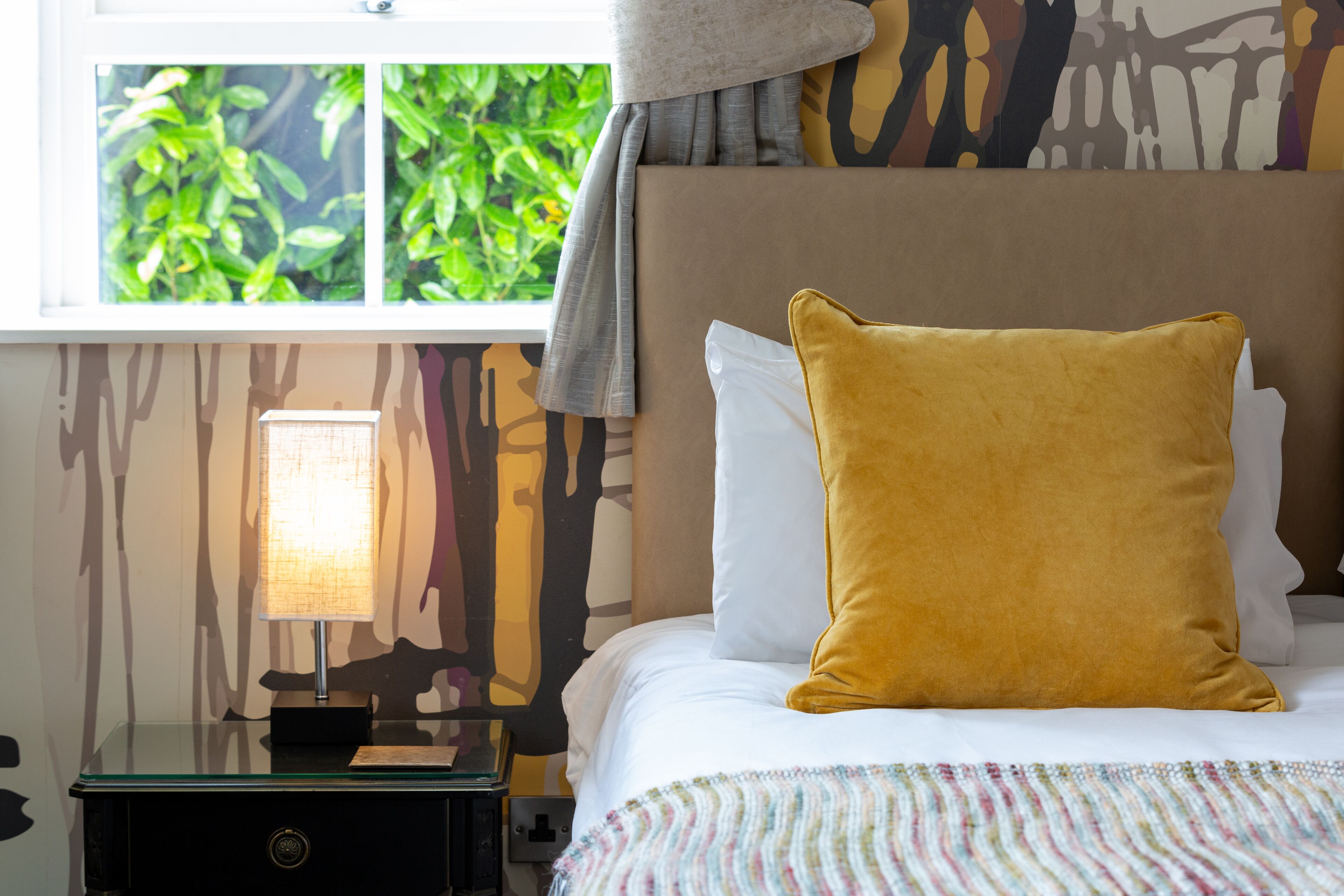 Uma cama bem arrumada com travesseiro amarelo e manta listrada, mesa de cabeceira com abajur e janela com vista para o verde.
