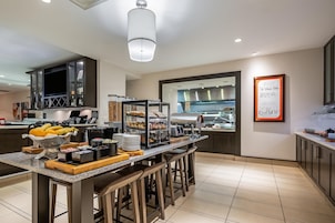 Um buffet de café da manhã moderno, com diversas opções de alimentos, área de estar com banquinhos e vista clara para a cozinha ao fundo.