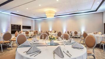 Salão de banquetes com mesas redondas preparadas para refeições, equipado com cadeiras, louças e um lustre central.
