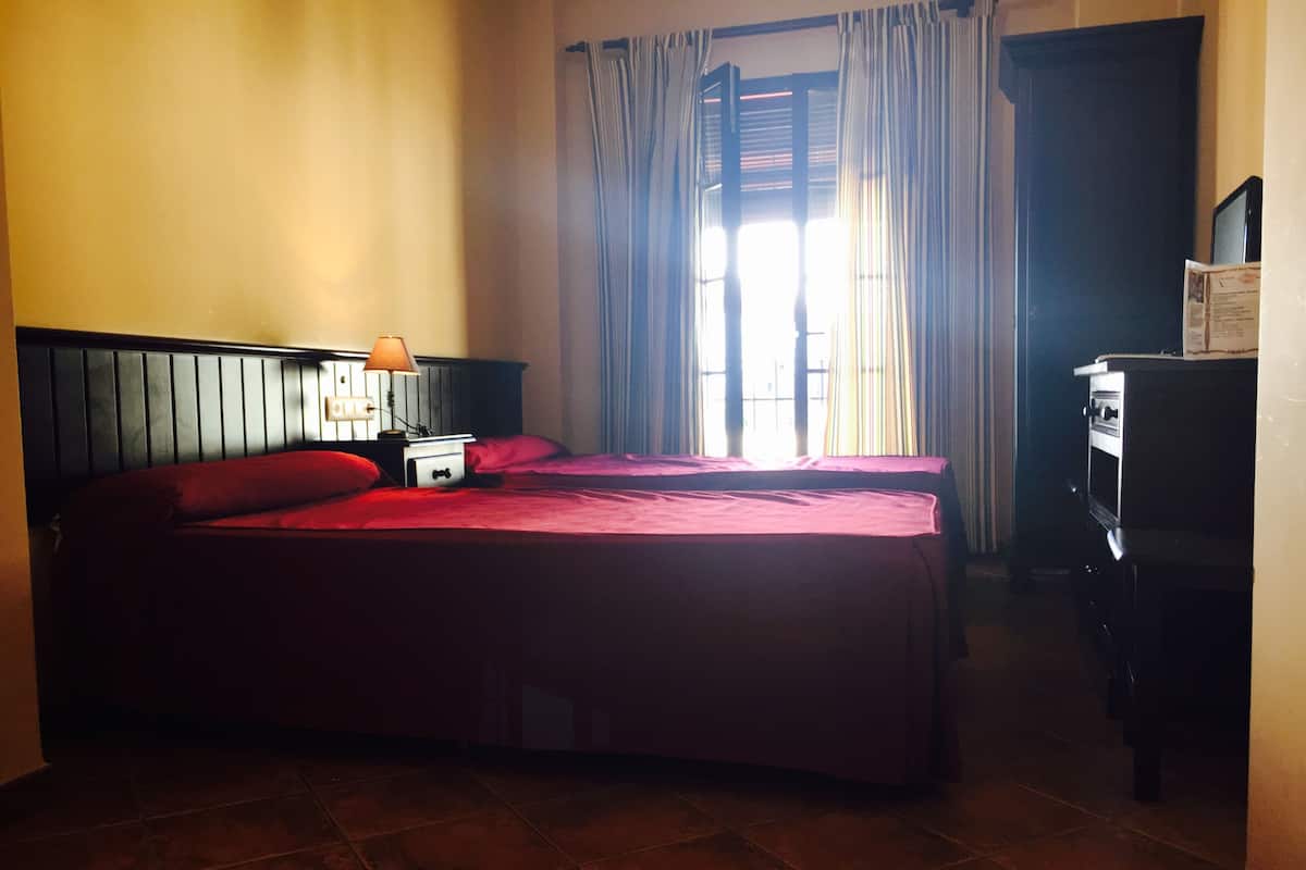Quarto de hotel com cama, mesa de cabeceira, abajur, cômoda e janela com cortinas.