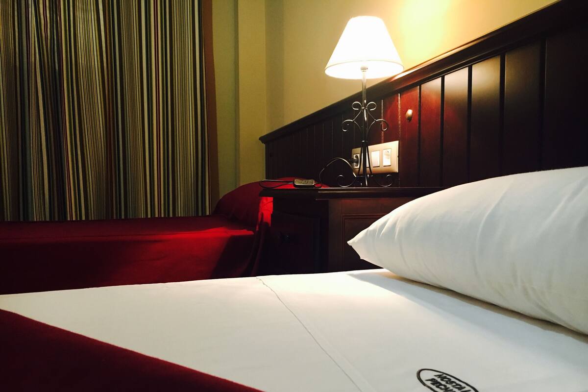 Quarto de hotel com cama, mesa de cabeceira, luminária e cortina.