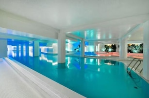 Uma piscina interior com água cristalina, rodeada por paredes e colunas brancas, e vista para o céu noturno.