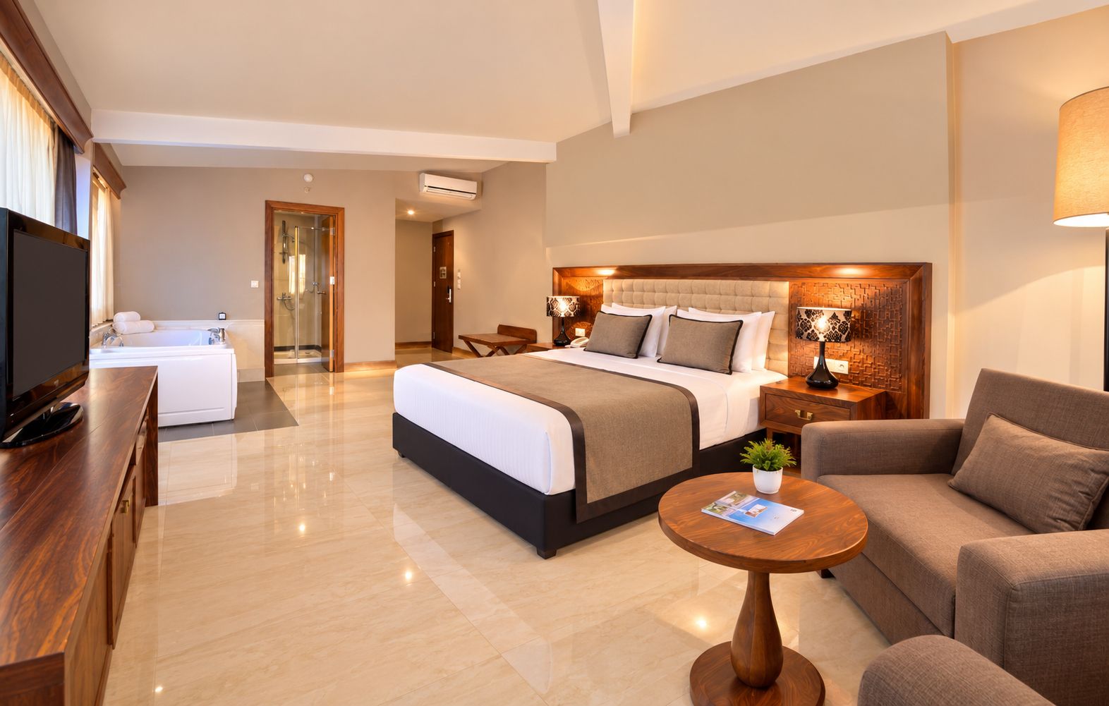 Quarto de hotel com cama, sofá, televisão, mesa de centro e banheiro visível ao fundo.