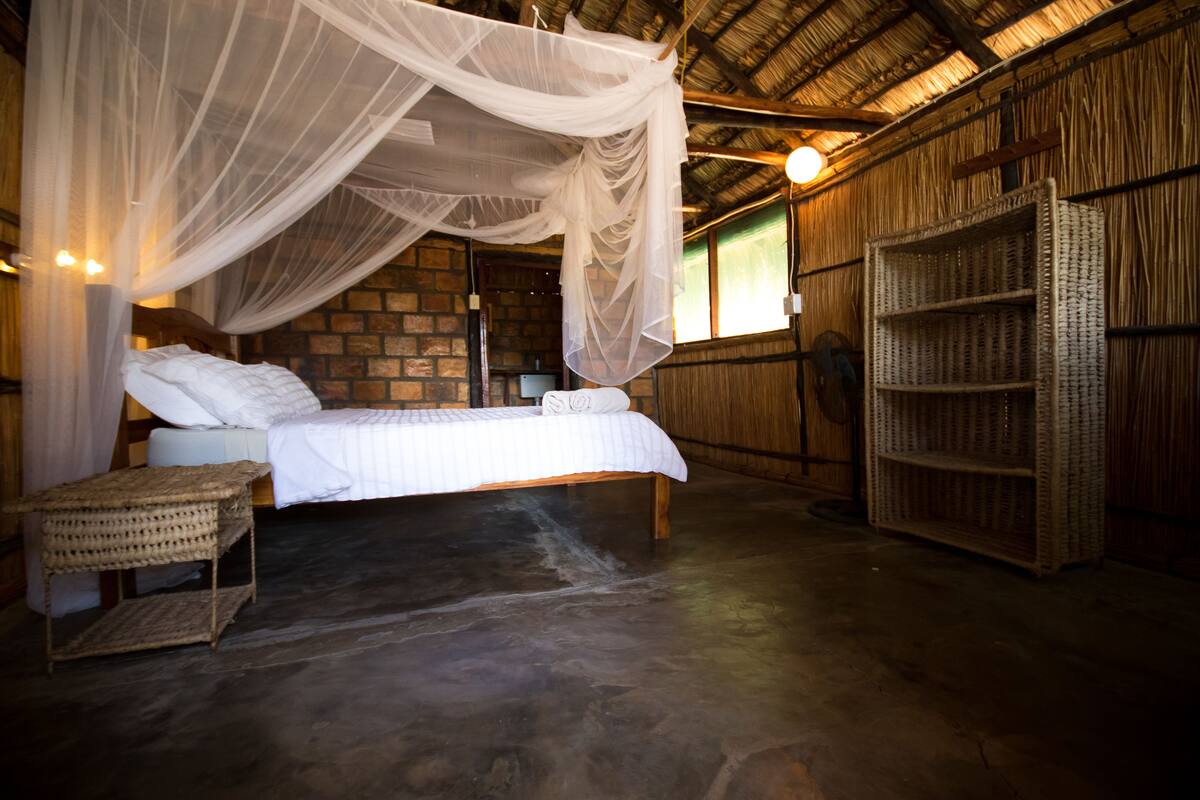 Quarto com uma cama, uma mesinha de vime, uma estante de tecido e uma rede mosquiteira.
