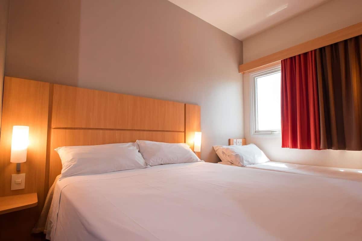 Quarto de hotel com uma cama grande, abajures, uma janela com cortinas e uma cabeceira de madeira.