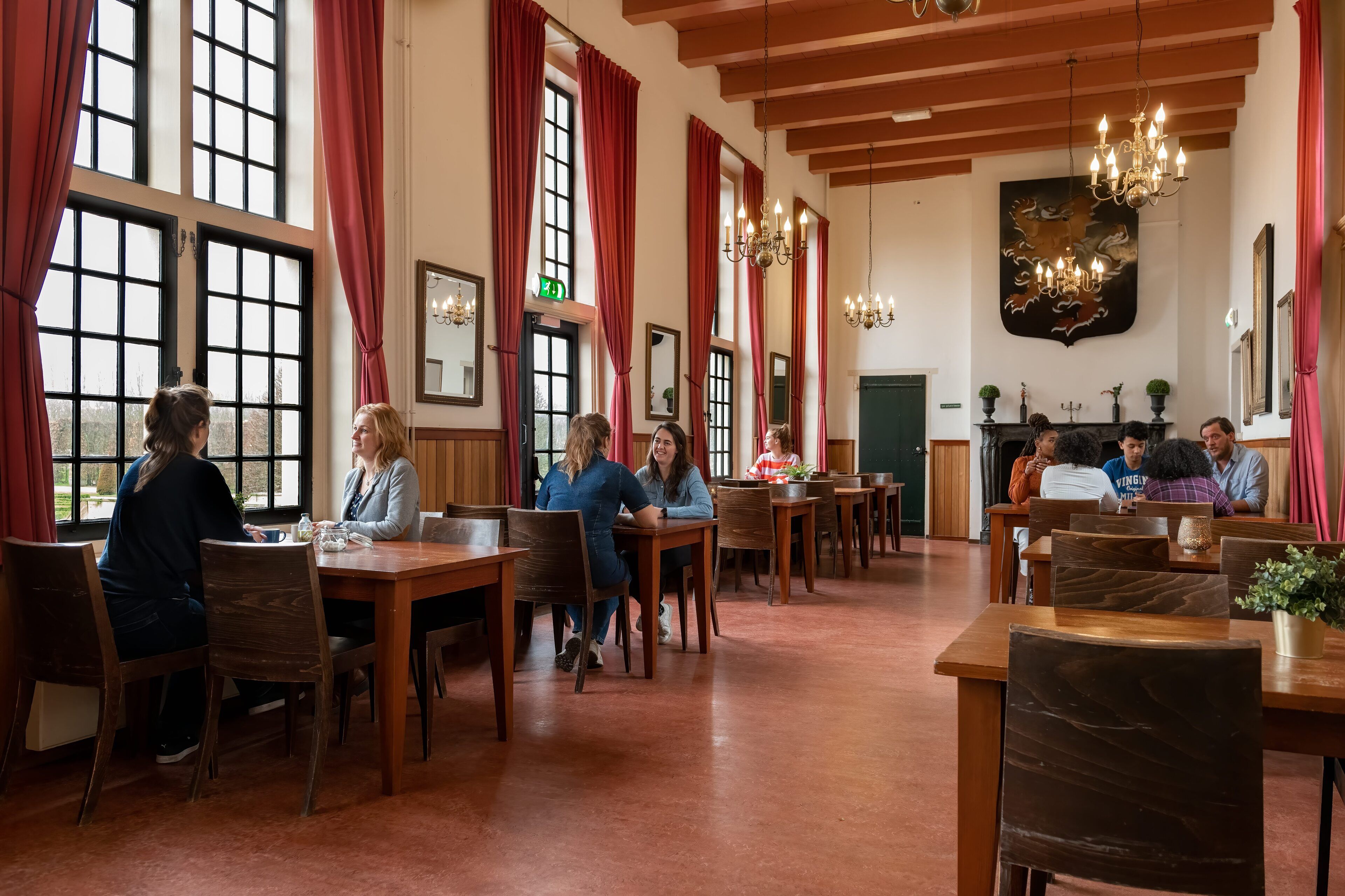 Uma sala de jantar espaçosa com mesas e cadeiras de madeira, grandes janelas com cortinas vermelhas e um candelabro.
