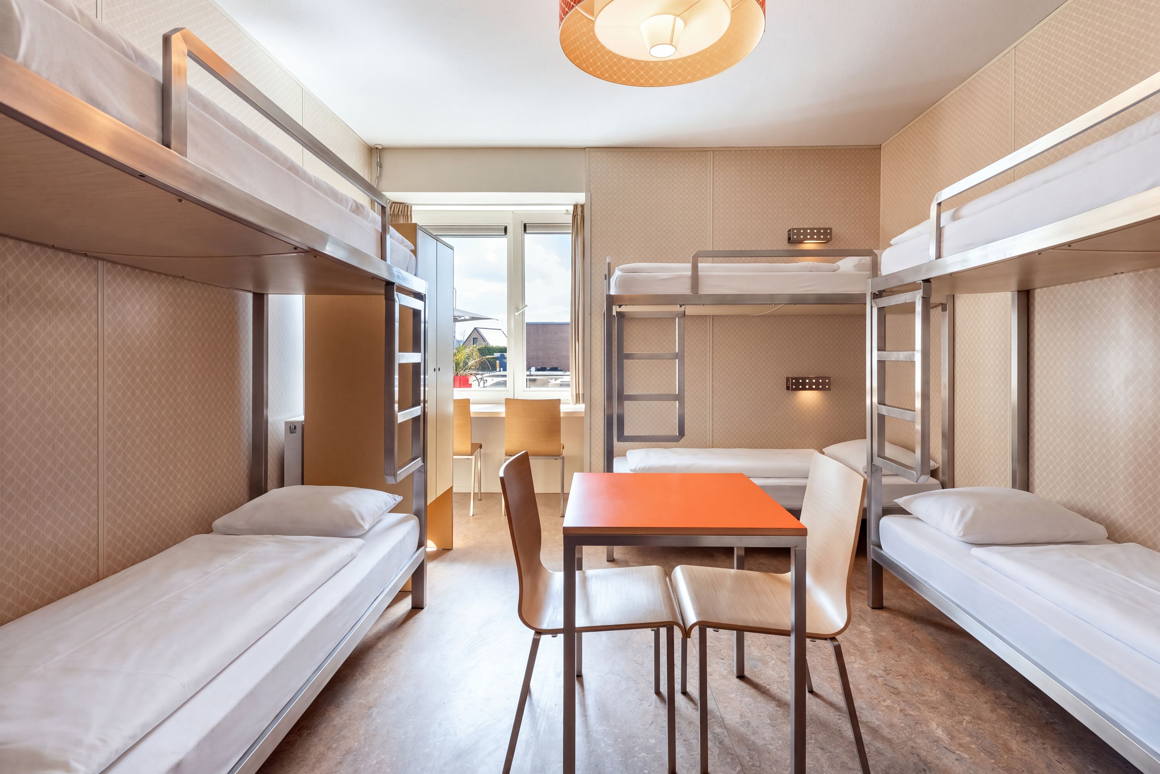 Quarto de hotel pequeno com beliches, uma mesinha e cadeiras.