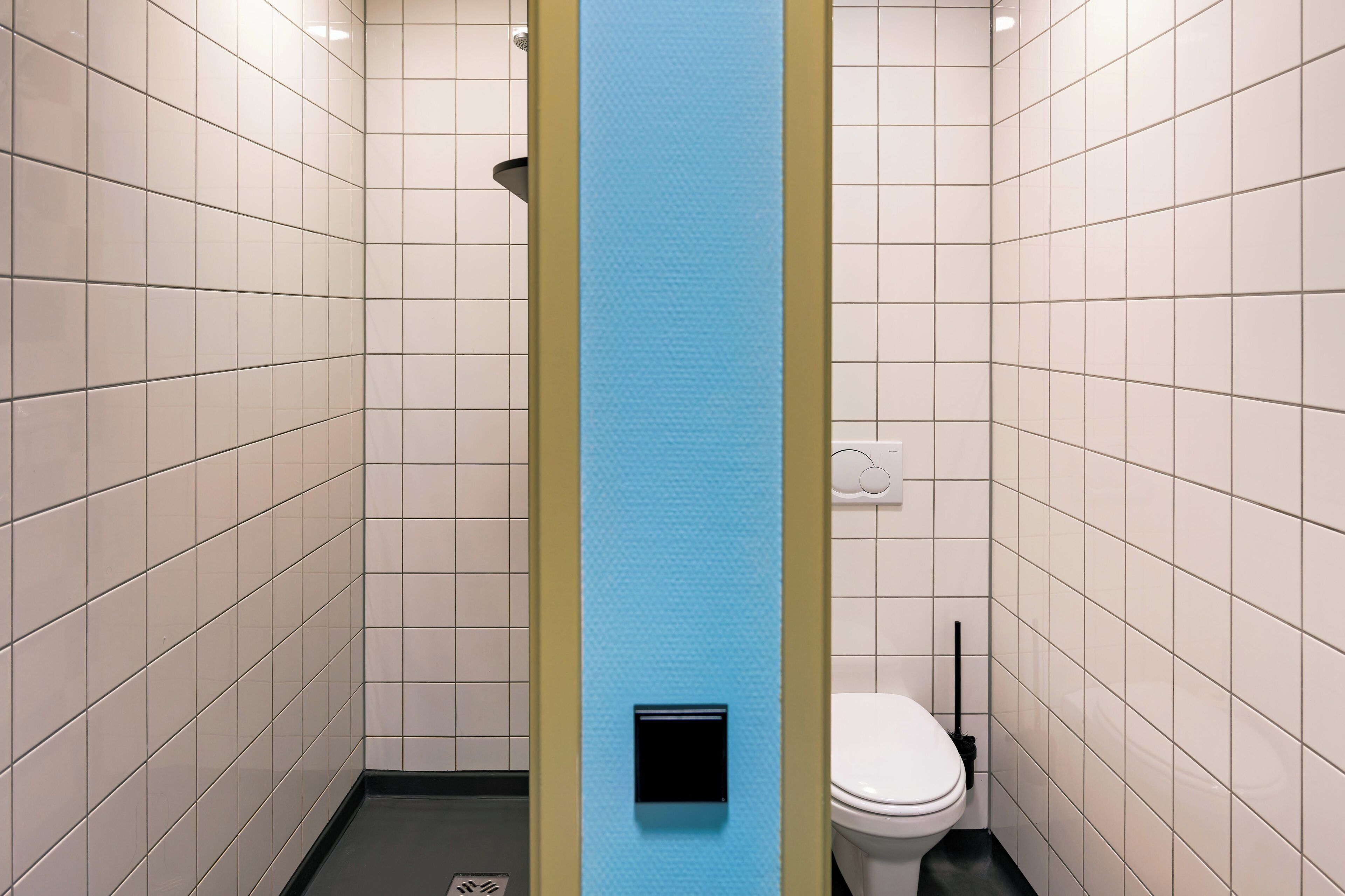 Casa de banho pequena, com azulejos, sanita branca, faixa vertical azul na parede e ralos de chão pretos.