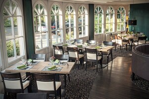 Um restaurante com grandes janelas em arco, mesas e cadeiras de madeira.