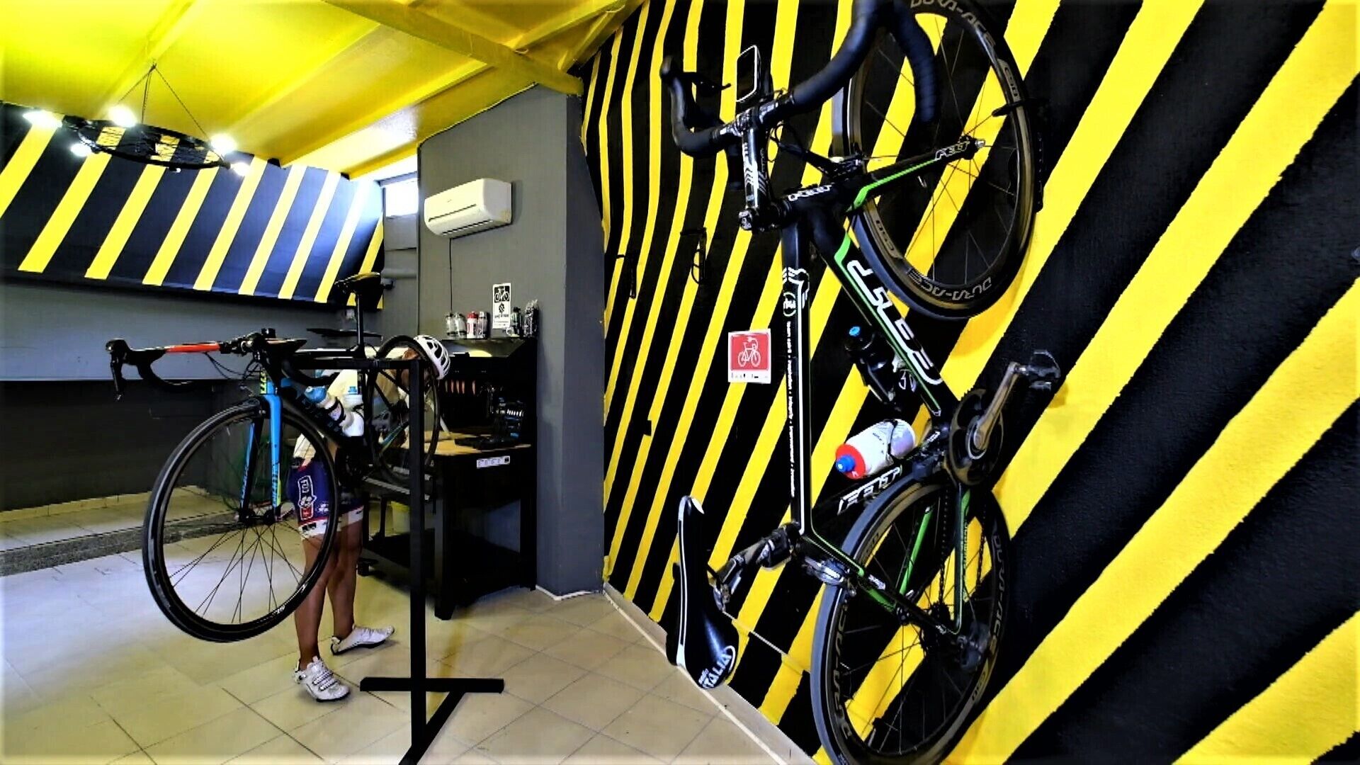 Quarto com bicicletas penduradas na parede, uma escrivaninha com um computador e uma pessoa ao lado de uma bicicleta.