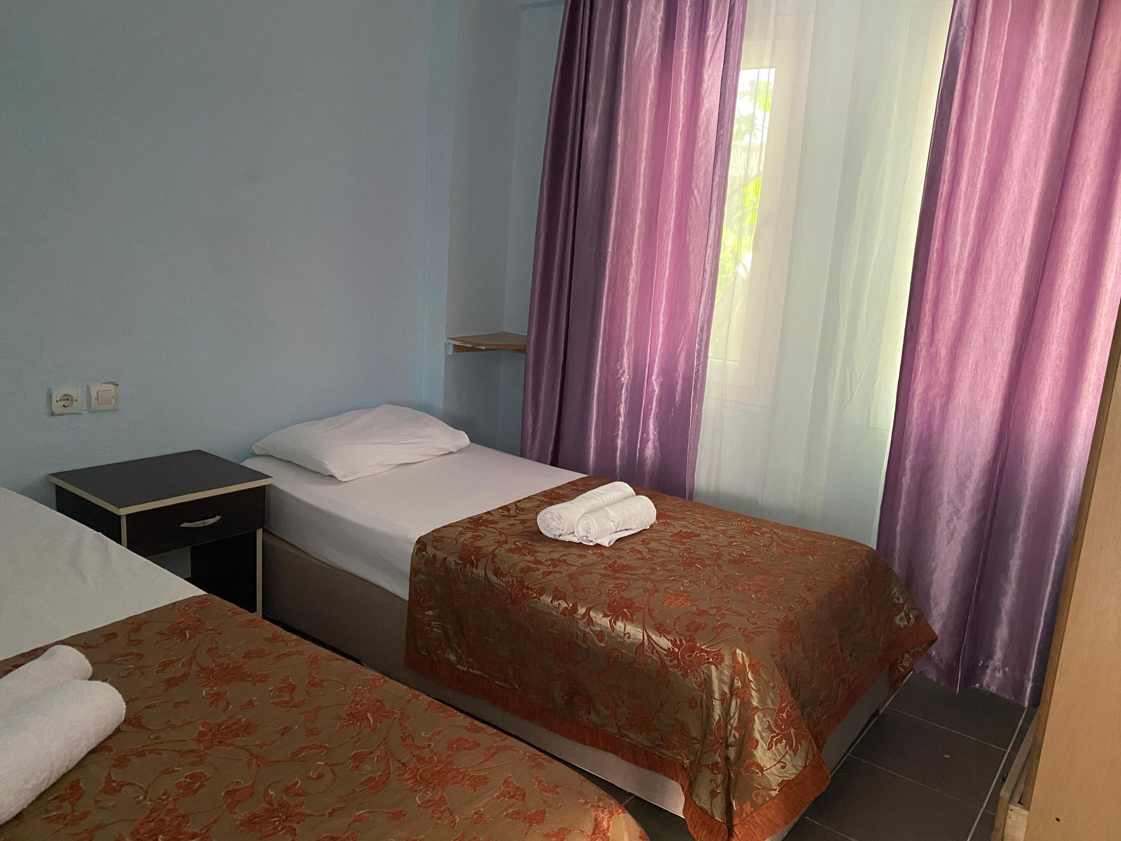 Quarto de hotel com duas camas, uma mesinha de cabeceira e uma janela com cortinas roxas.