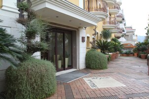Entrada de hotel com caminho de tijolos, plantas em vasos e tapete de boas-vindas com o nome do hotel.