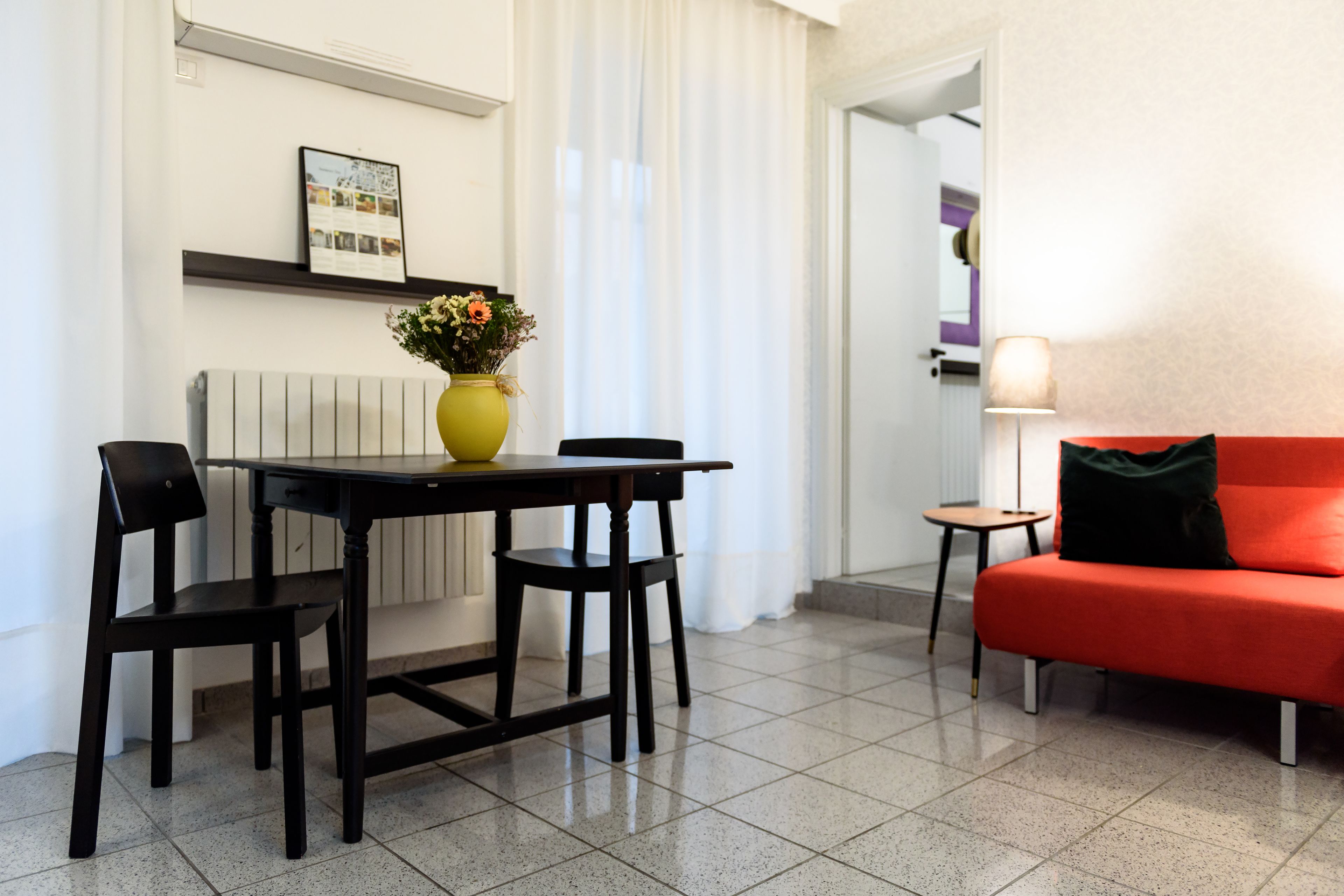 Uma sala de estar moderna com área de jantar, apresentando um sofá vermelho, uma mesa pequena com cadeiras, um vaso amarelo com flores e uma moldura na parede.