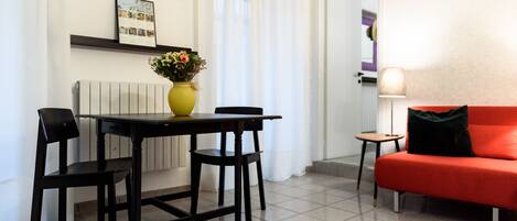 Uma sala de estar moderna com área de jantar, apresentando um sofá vermelho, uma mesa pequena com cadeiras, um vaso amarelo com flores e uma moldura na parede.