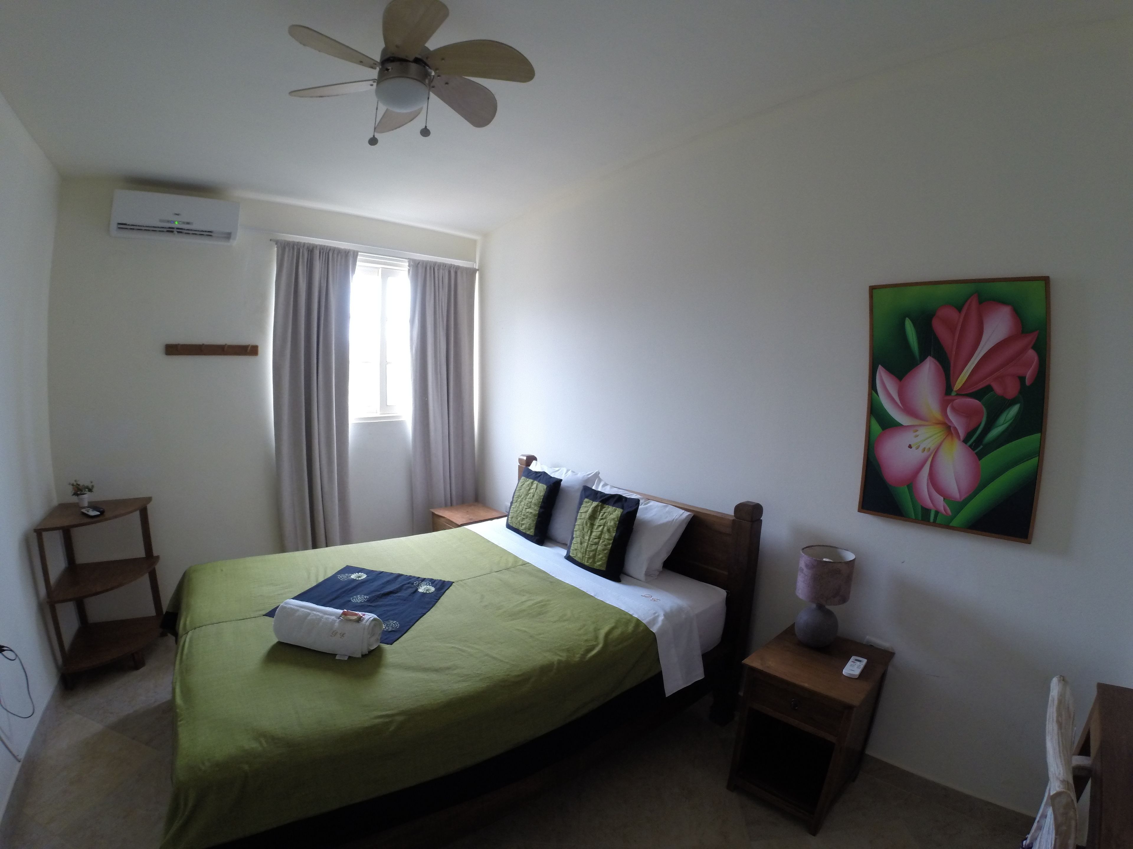 Quarto de hotel com uma cama de solteiro, mesa de cabeceira, ventoinha no teto, ar condicionado, janela com cortinas e um quadro com flores na parede.