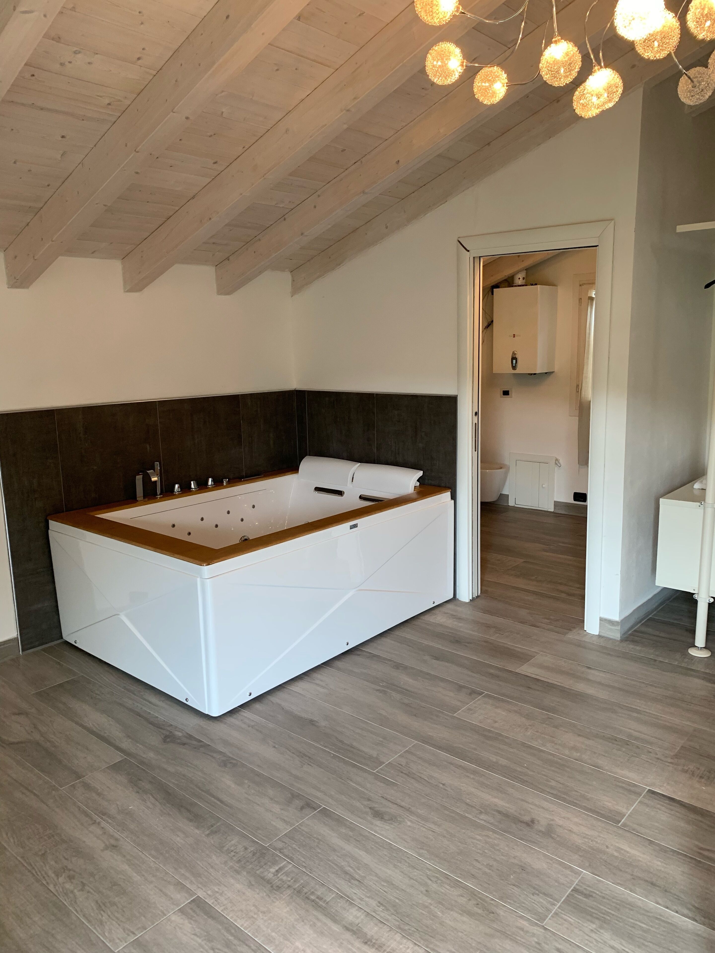 Um quarto moderno com uma cama grande, estrutura branca em madeira, cabeceira escura e piso de madeira. O teto possui vigas aparentes e um lustre.