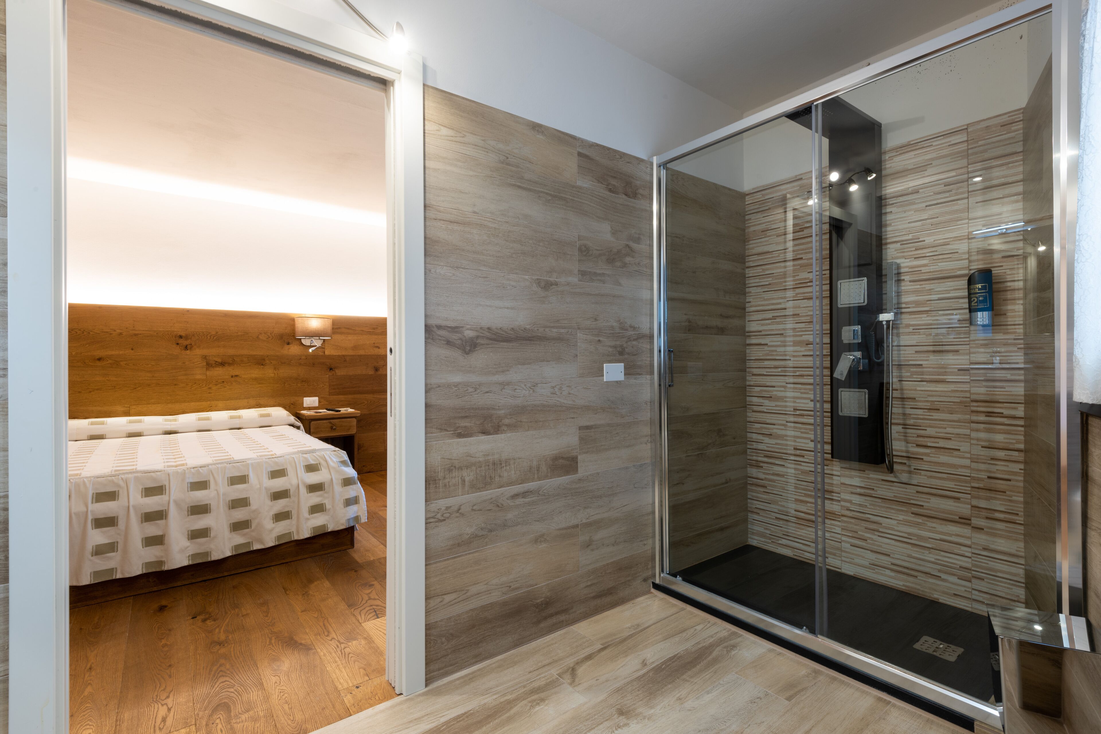 Quarto moderno com piso de madeira, cama com colcha estampada, mesa de cabeceira e box de vidro.