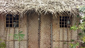 Uma cabana com telhado de palha, três janelas, uma porta central e parede trançada.