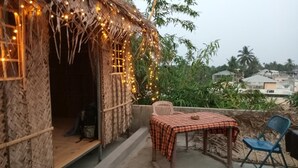Uma construção com telhado de palha, luzes de corda, uma mesa xadrez e cadeiras em uma varanda com vista para edifícios e vegetação ao redor.