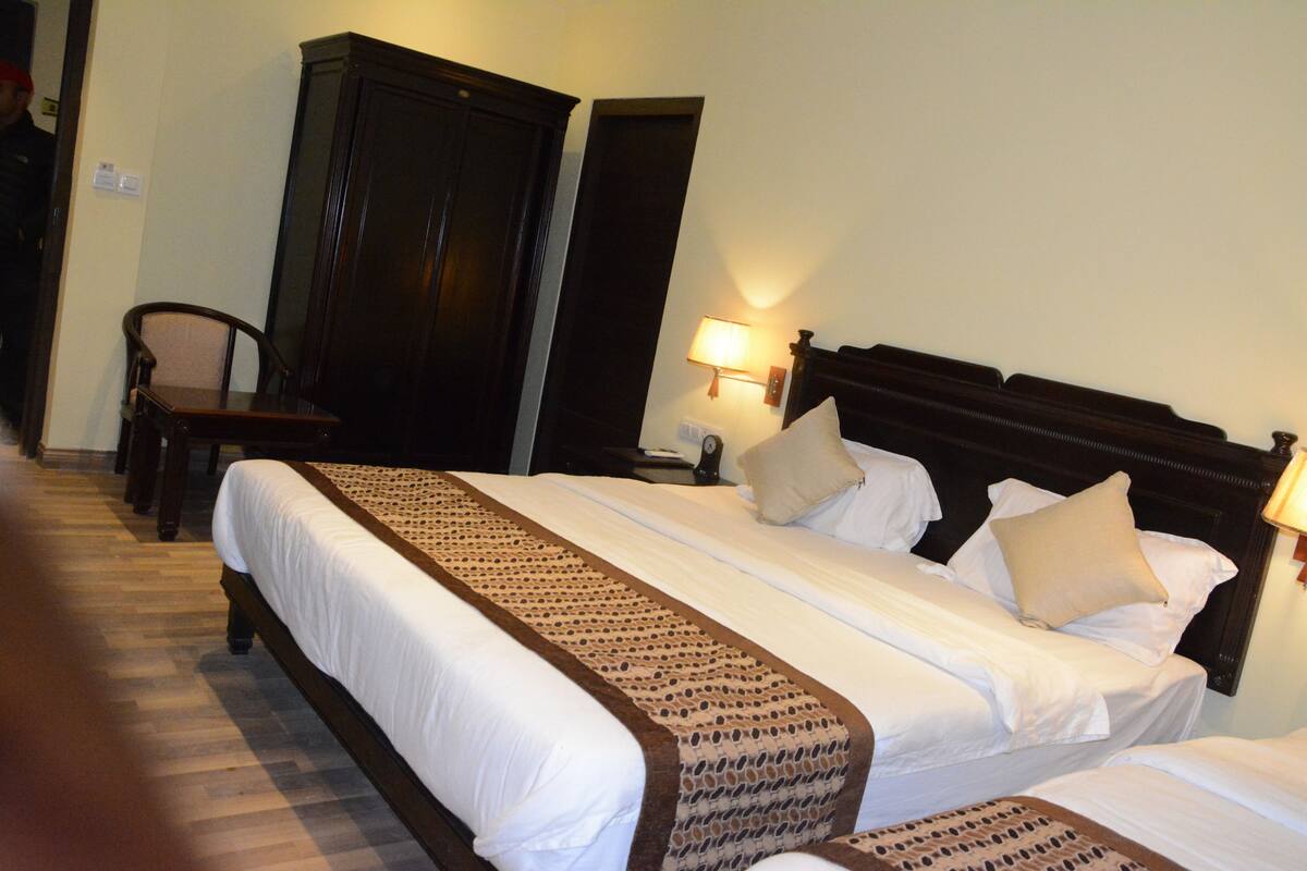 Quarto de hotel com duas camas, guarda-roupa de madeira, uma cadeira e mesa de cabeceira com abajur.